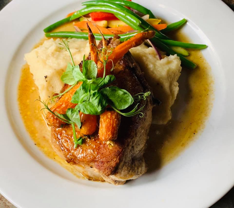 A plate of elegantly plated food, featuring a seared pork chop, creamy mashed potatoes, a selection of roasted carrots and green beans, all drizzled with a savory gravy.