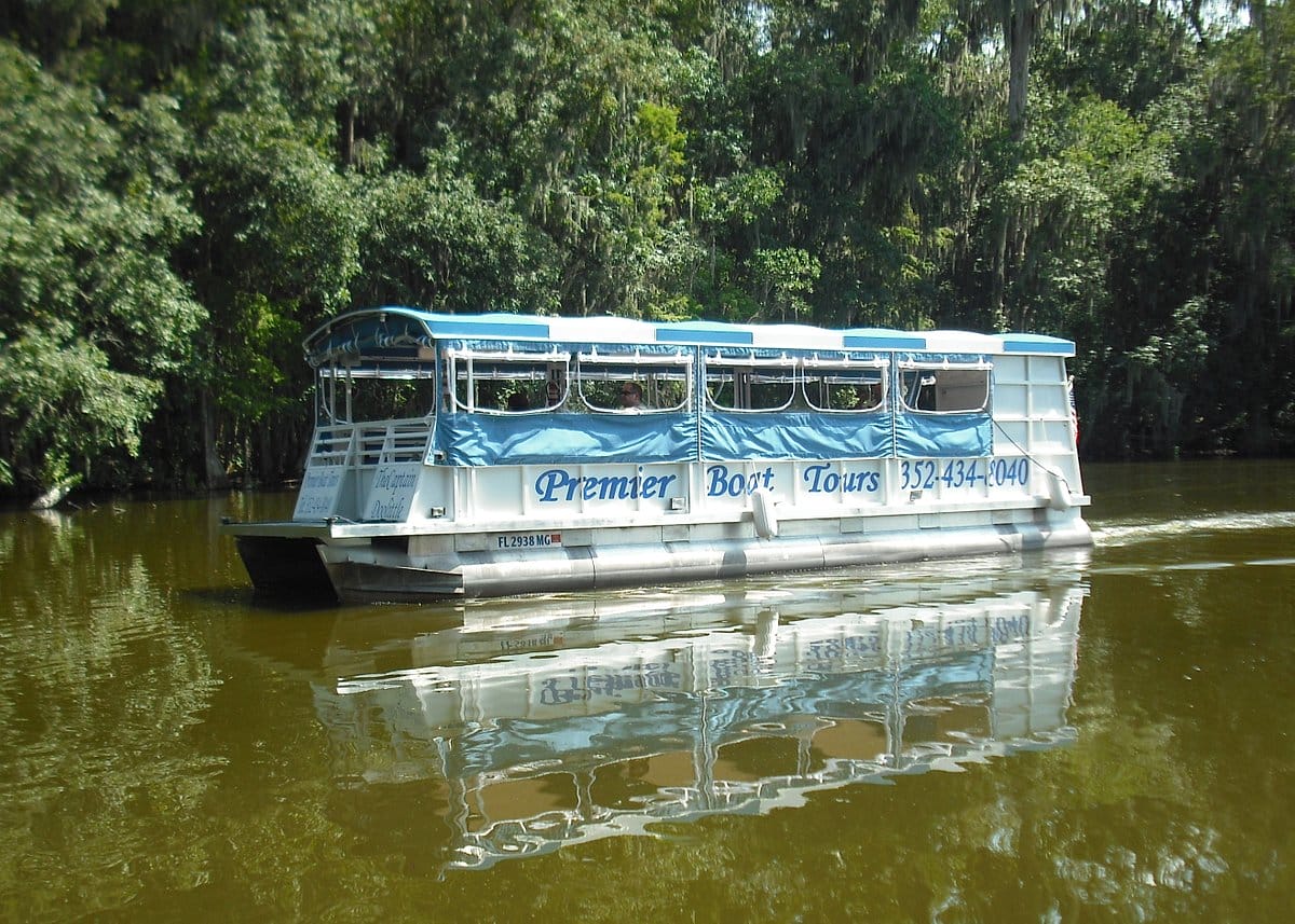 A covered tour boat named "Premier Boat Tours" floats on a calm river, surrounded by dense greenery reflecting on the water's surface.