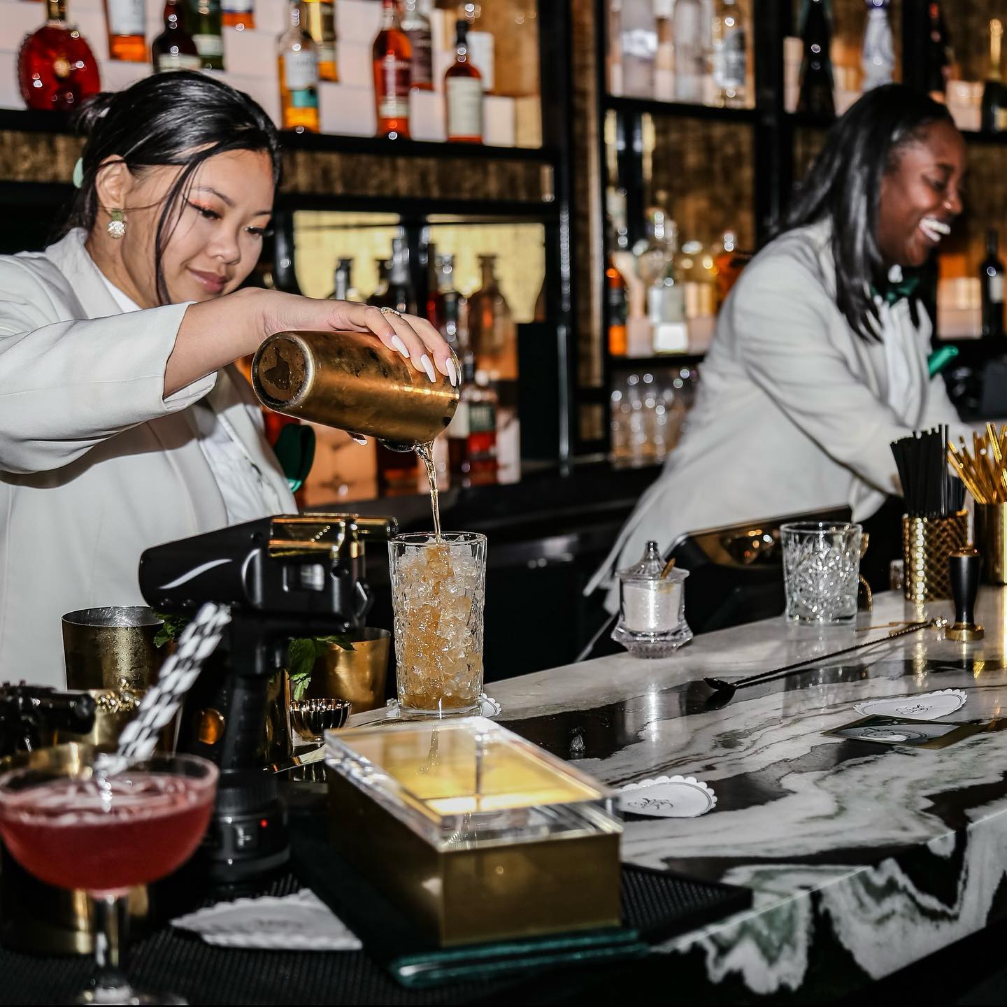 a bartender in a white coat pouring a drink behind a marble bar with a gleaming backdrop and a smiling colleague