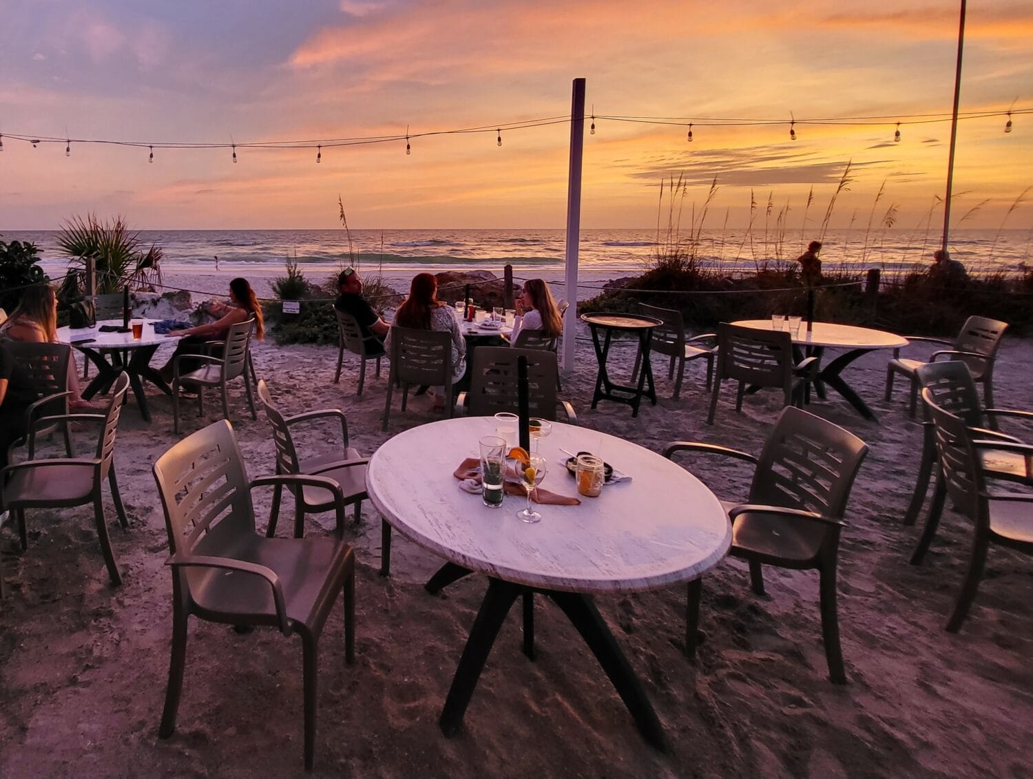 a beachfront dining setup with tables and chairs on the sand overlooking the sea at sunset