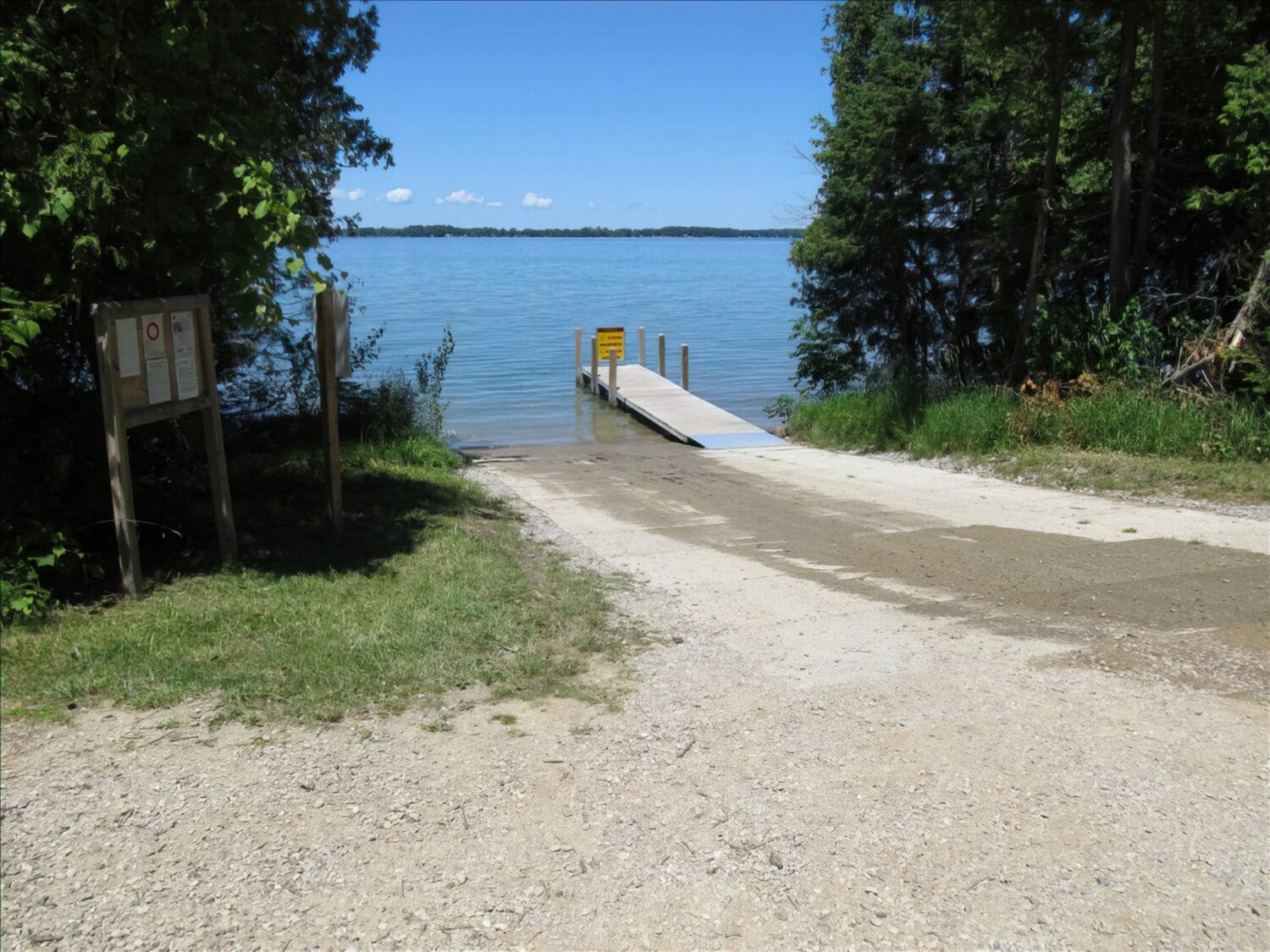 a boat ramp leading into a clear lake flanked by greenery and a notice board on the left