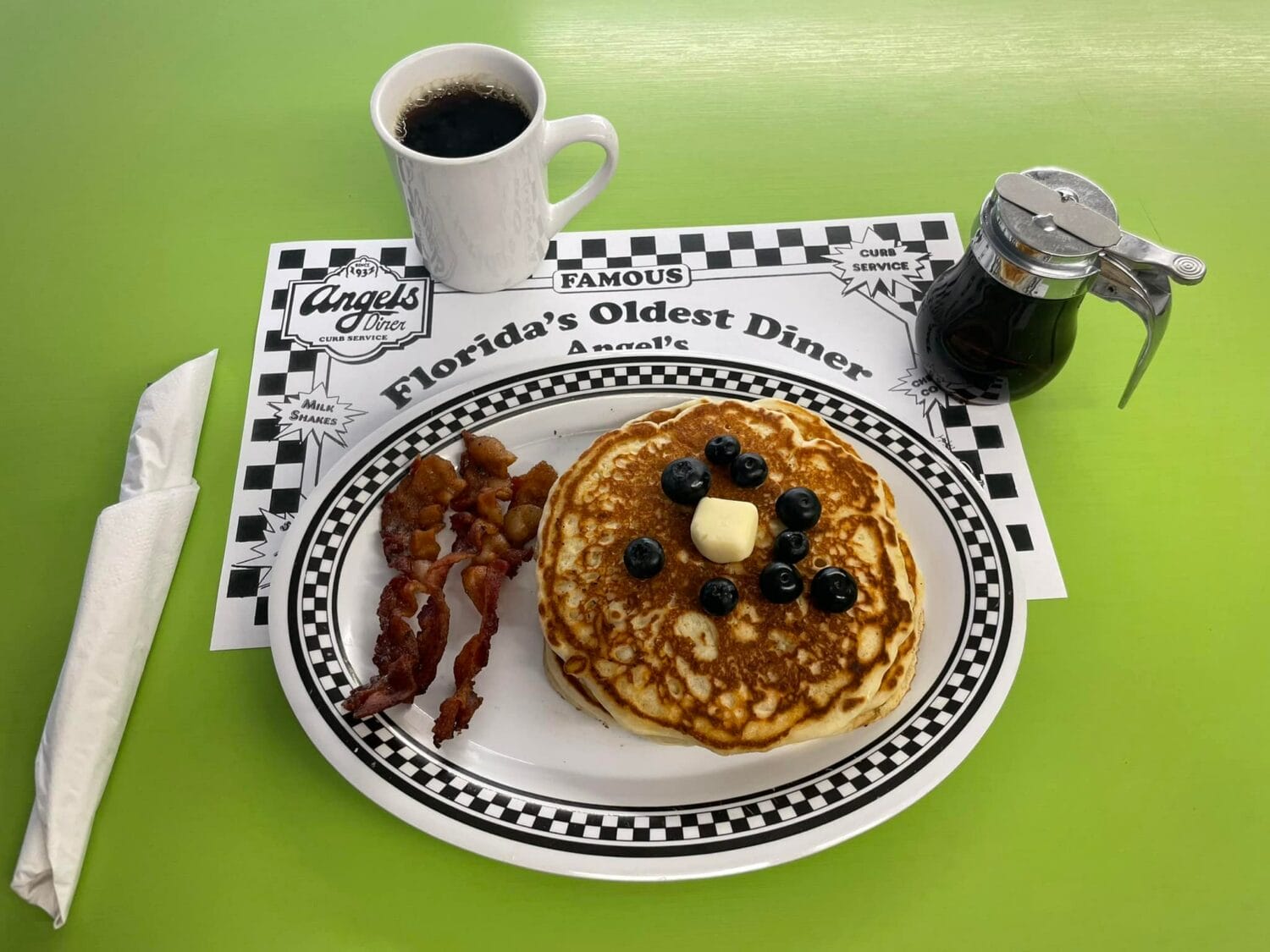 a breakfast plate with pancakes topped with butter and blueberries bacon strips and a cup of coffee on a vibrant green countertop