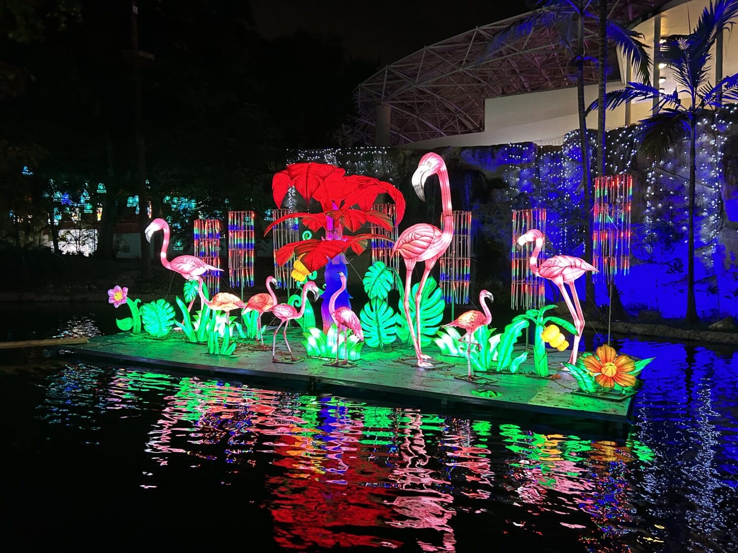 a colorful illuminated display of flamingos and tropical flowers on a water platform with reflections creating a dazzling effect on the water surface at night