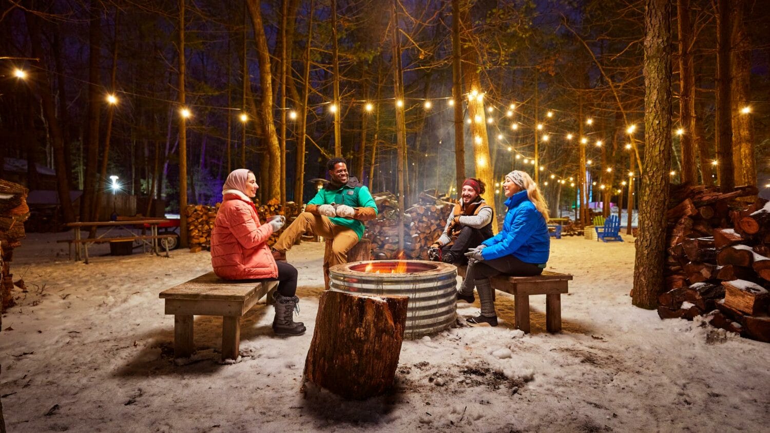 A group of friends enjoying a convivial moment around a blazing fire pit in a snowy forest, illuminated by string lights in the evening.