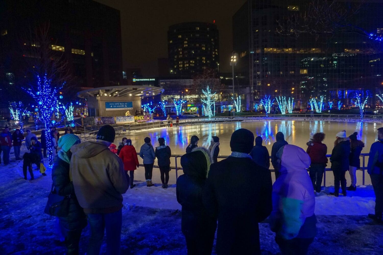 a group of onlookers watches skaters glide across the ice rink on a cold evening with the city illuminated by festive lights