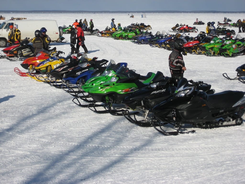 a line of colorful snowmobiles parked on a snowy field with riders gathered around