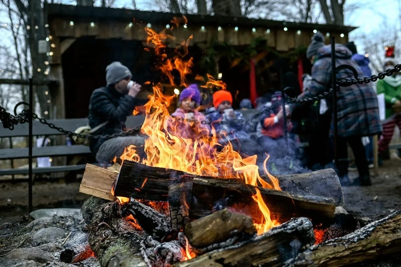 a lively bonfire blazes in the foreground as people in winter attire enjoy drinks and conversations in a rustic outdoor setting