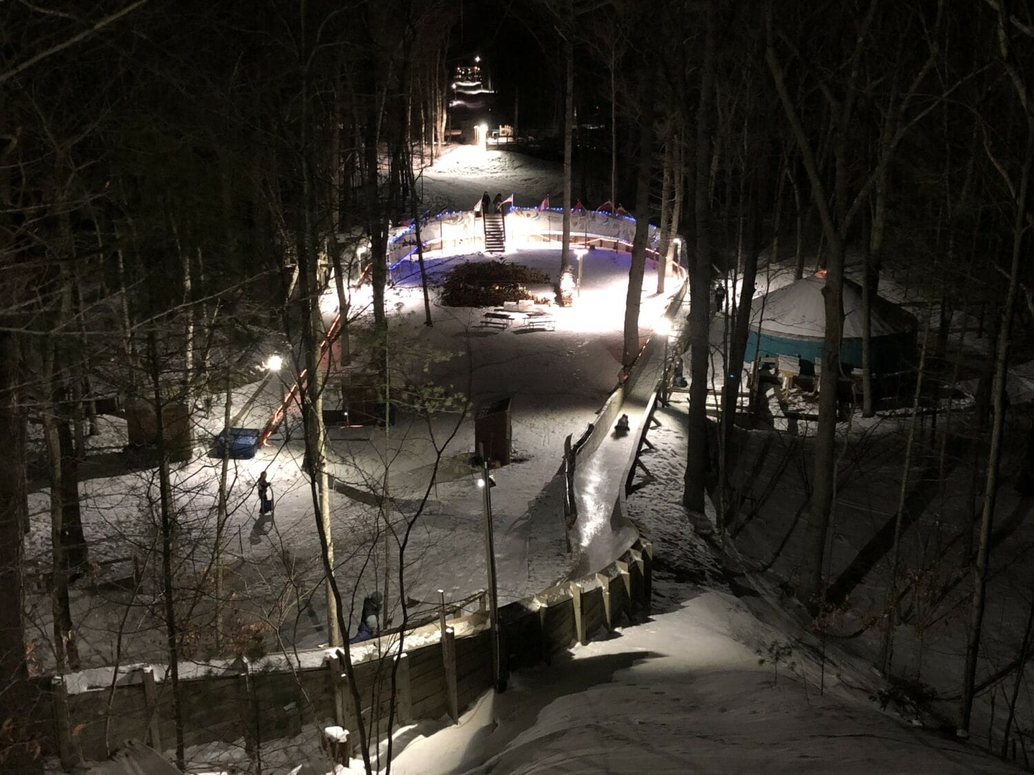 a nighttime view of a well lit outdoor luge track in a snowy forest setting with people gathered around a warm fire pit