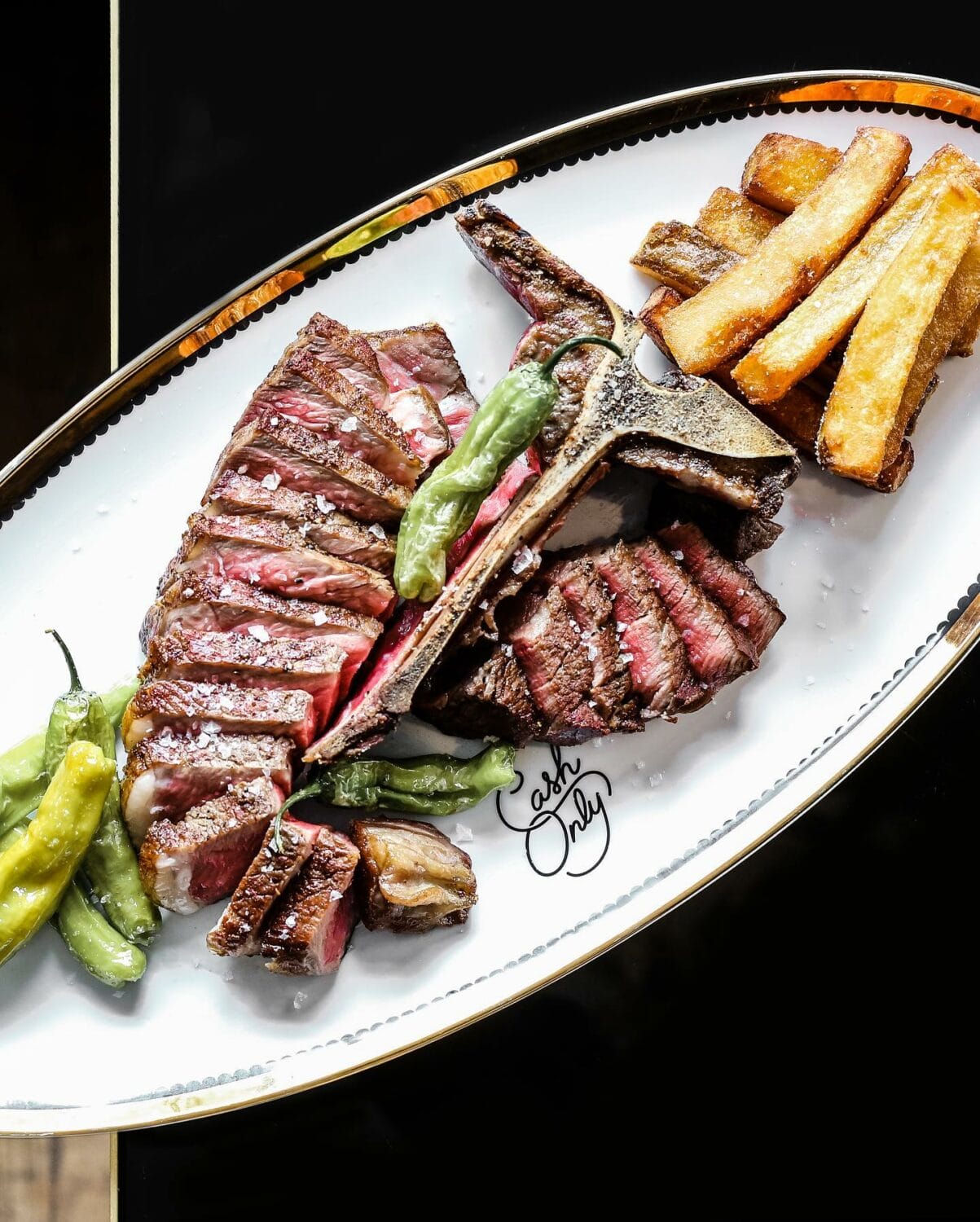 a plate of perfectly cooked steak sliced and served with golden fries and grilled shishito peppers on a white plate with a gold rim