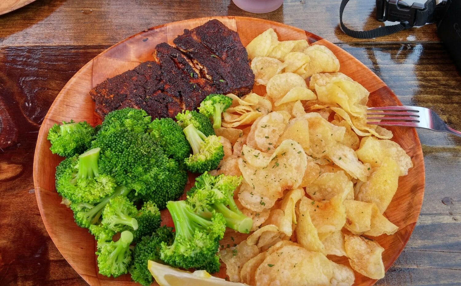a plate with blackened fish a side of broccoli and chips served with a lemon wedge on a wooden table