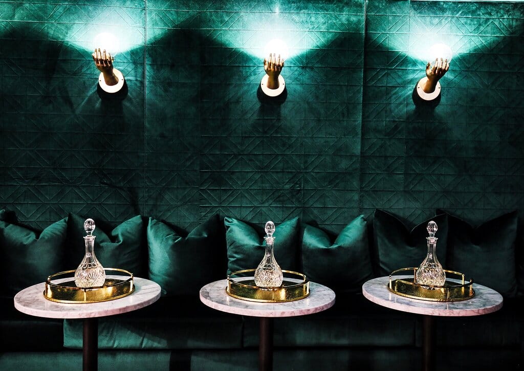 A posh lounge area with emerald green velvet pillows on a bench against a geometric-patterned wall, lit by unique hand-shaped sconces, with crystal decanters on marble side tables.