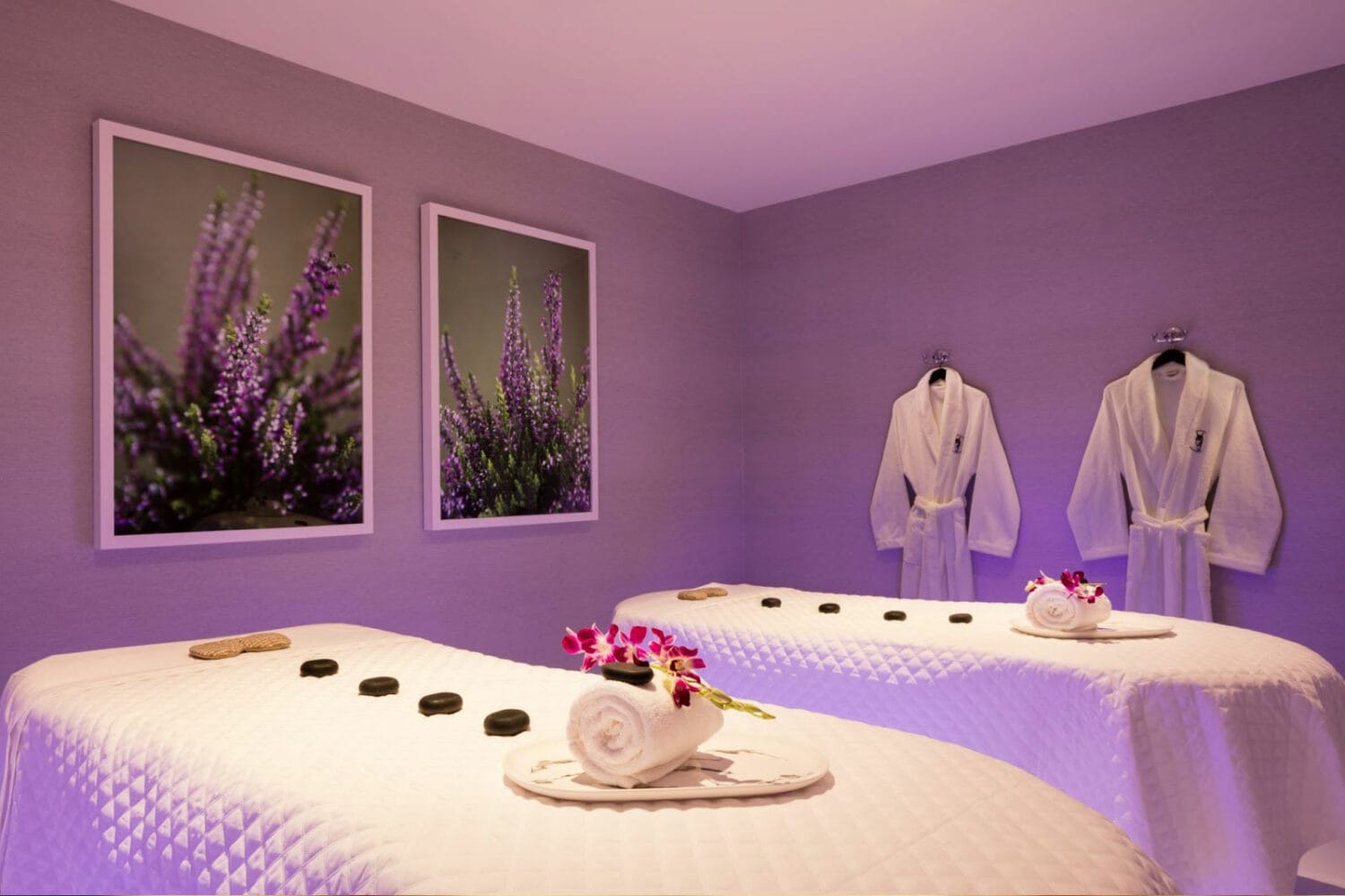 a serene spa room with twin massage tables soft purple lighting and framed pictures of lavender on the wall