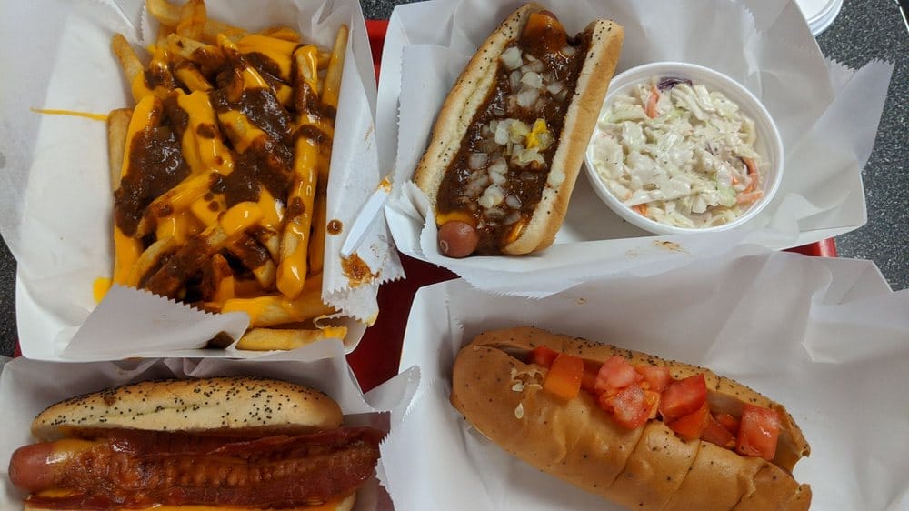 a variety of hot dogs with different toppings alongside french fries and onion rings on a gray countertop