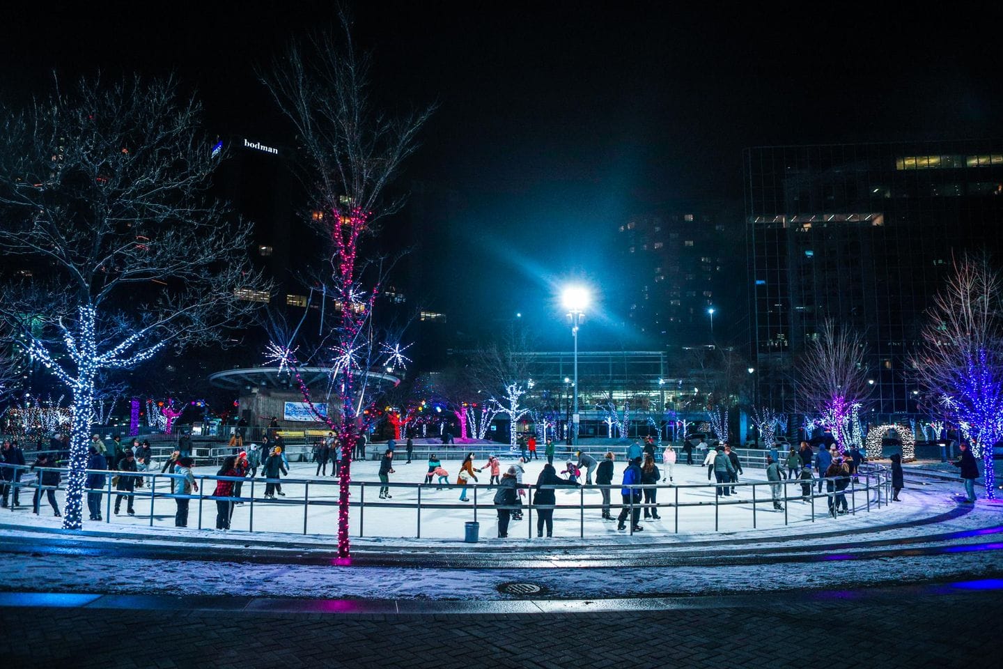 a vibrant nighttime scene at the ice skating rink with skaters surrounded by trees decorated with colorful lights