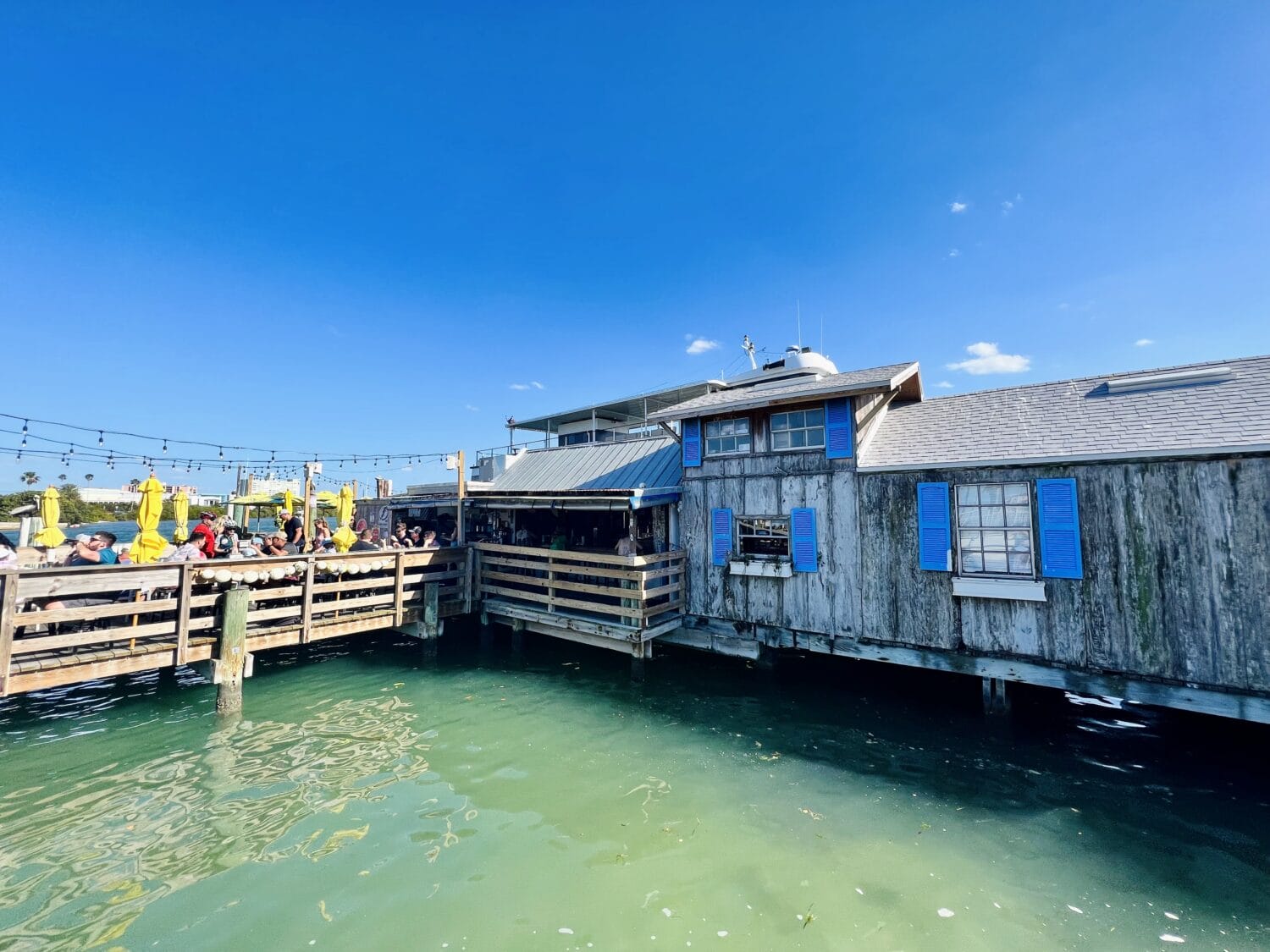 a waterfront dining area at a rustic restaurant with blue shutters where guests enjoy meals under yellow umbrellas on a sunny day