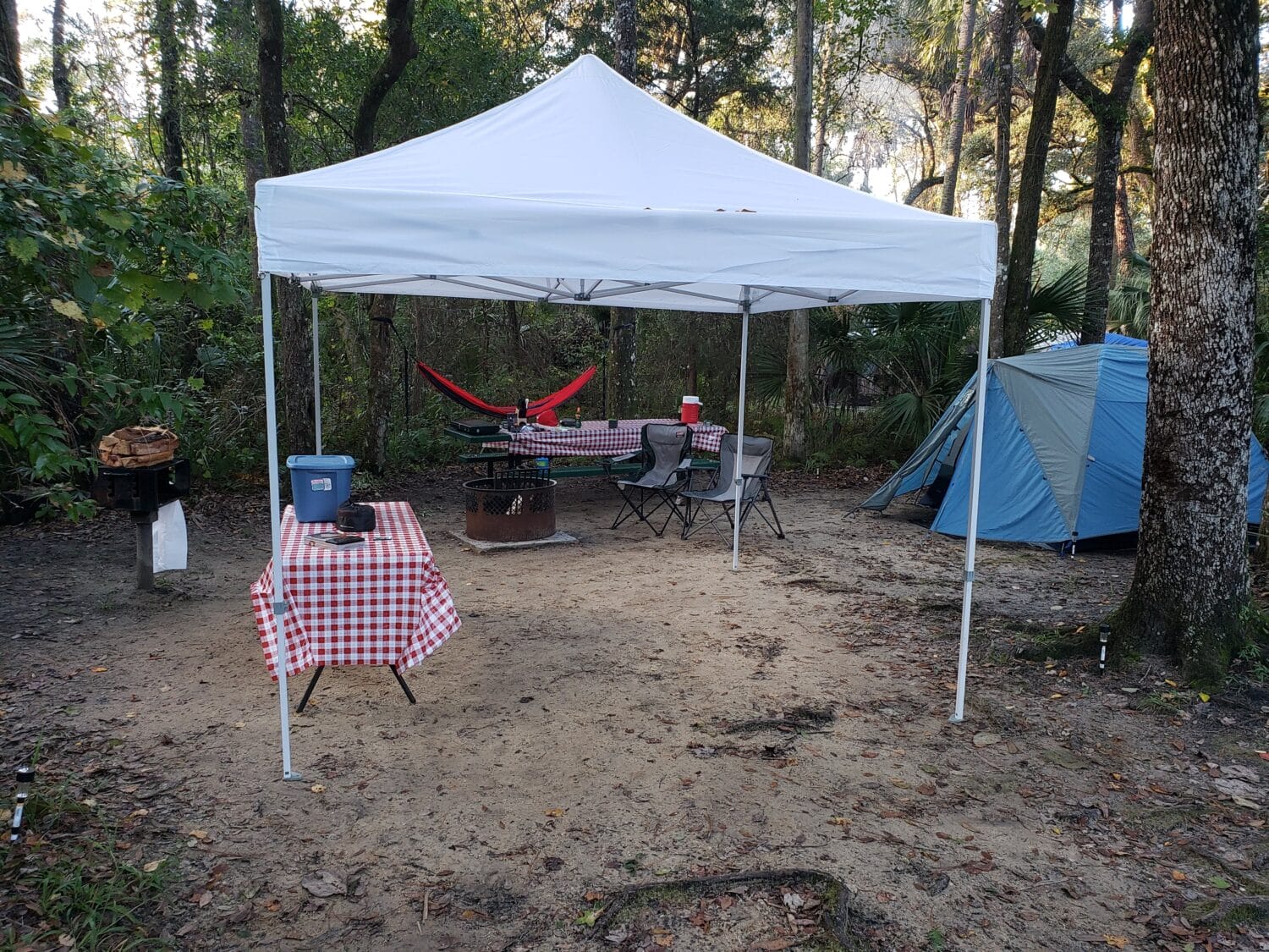 a well equipped campsite setup with a canopy a hammock a picnic table with a checkered tablecloth a portable grill and a tent in a forested area