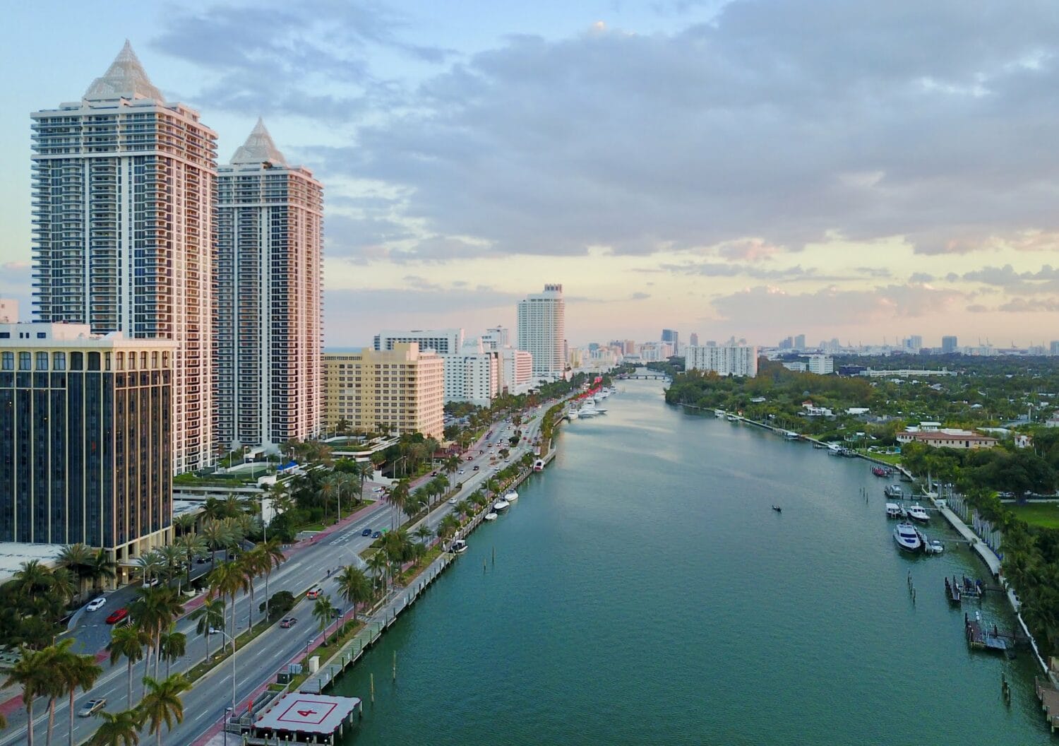 Aerial view of a sunlit urban waterfront in Florida lined with palm trees and tall buildings, with a clear view down a busy street