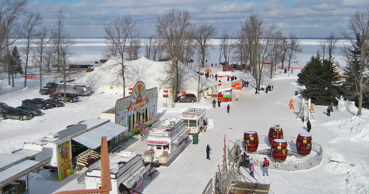 An Aerial view of Tip Up Town USA festival set against a backdrop of the frozen Houghton Lake, with visitors milling around food stands, rides, and attractions