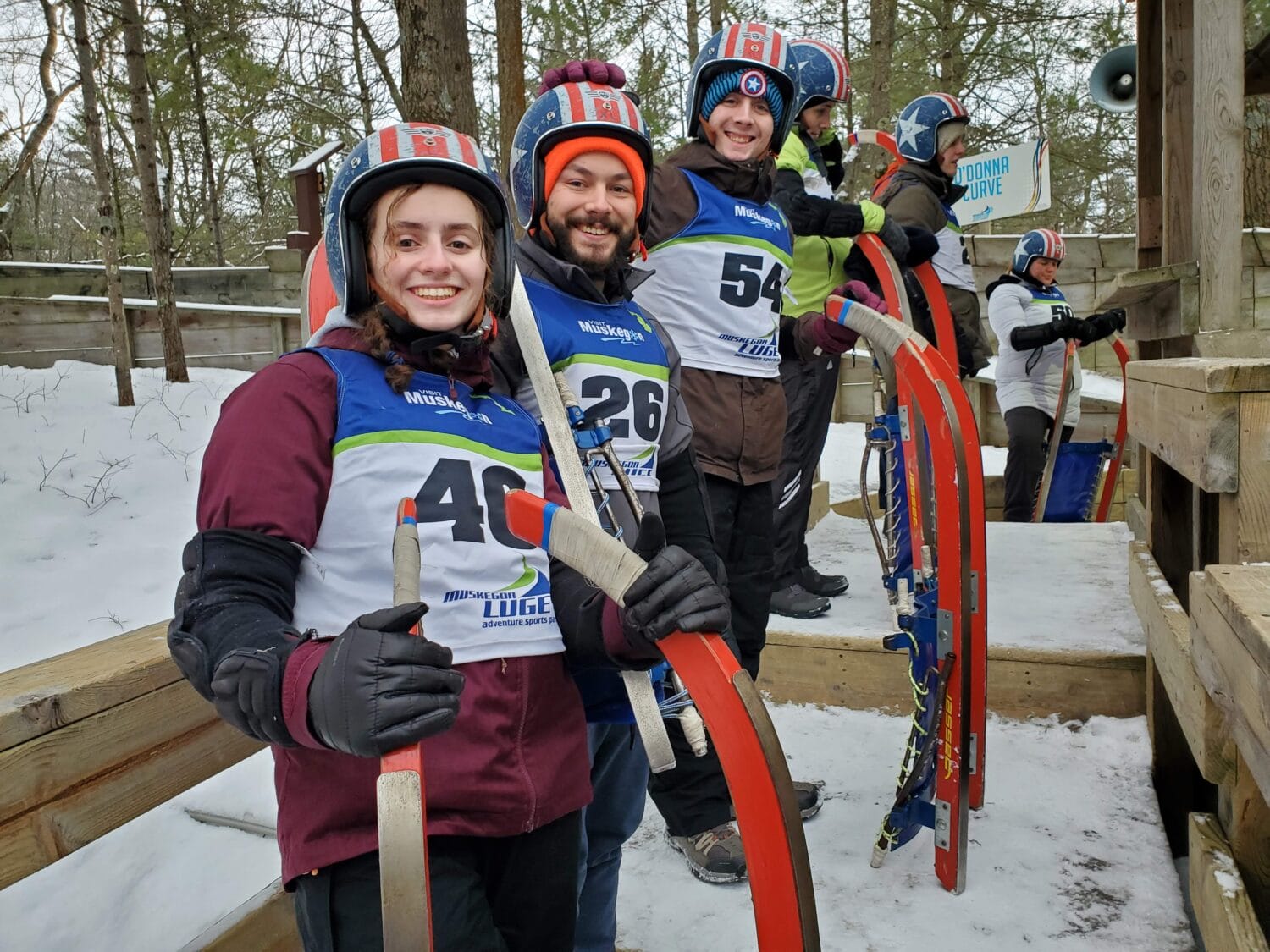 excited luge participants in colorful winter gear holding their sleds ready to descend the track with the starting area and forest backdrop