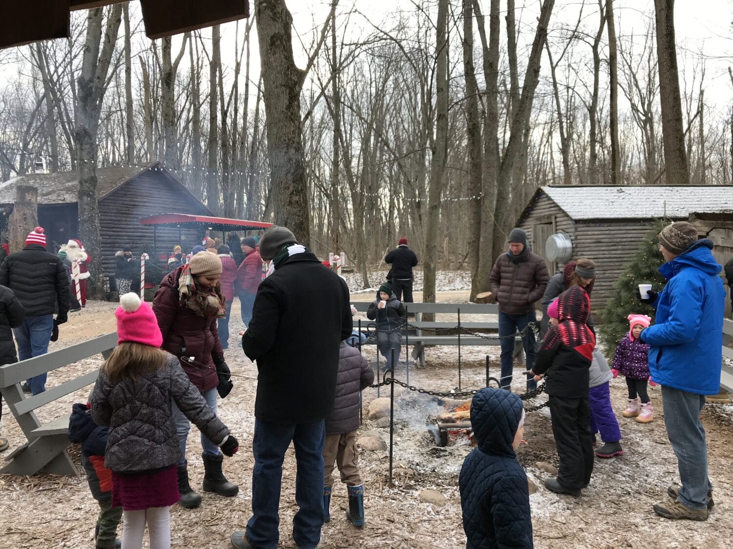 families and friends gather around picnic tables in a cozy winter clad forest clearing with a festive atmosphere and santa claus in the background