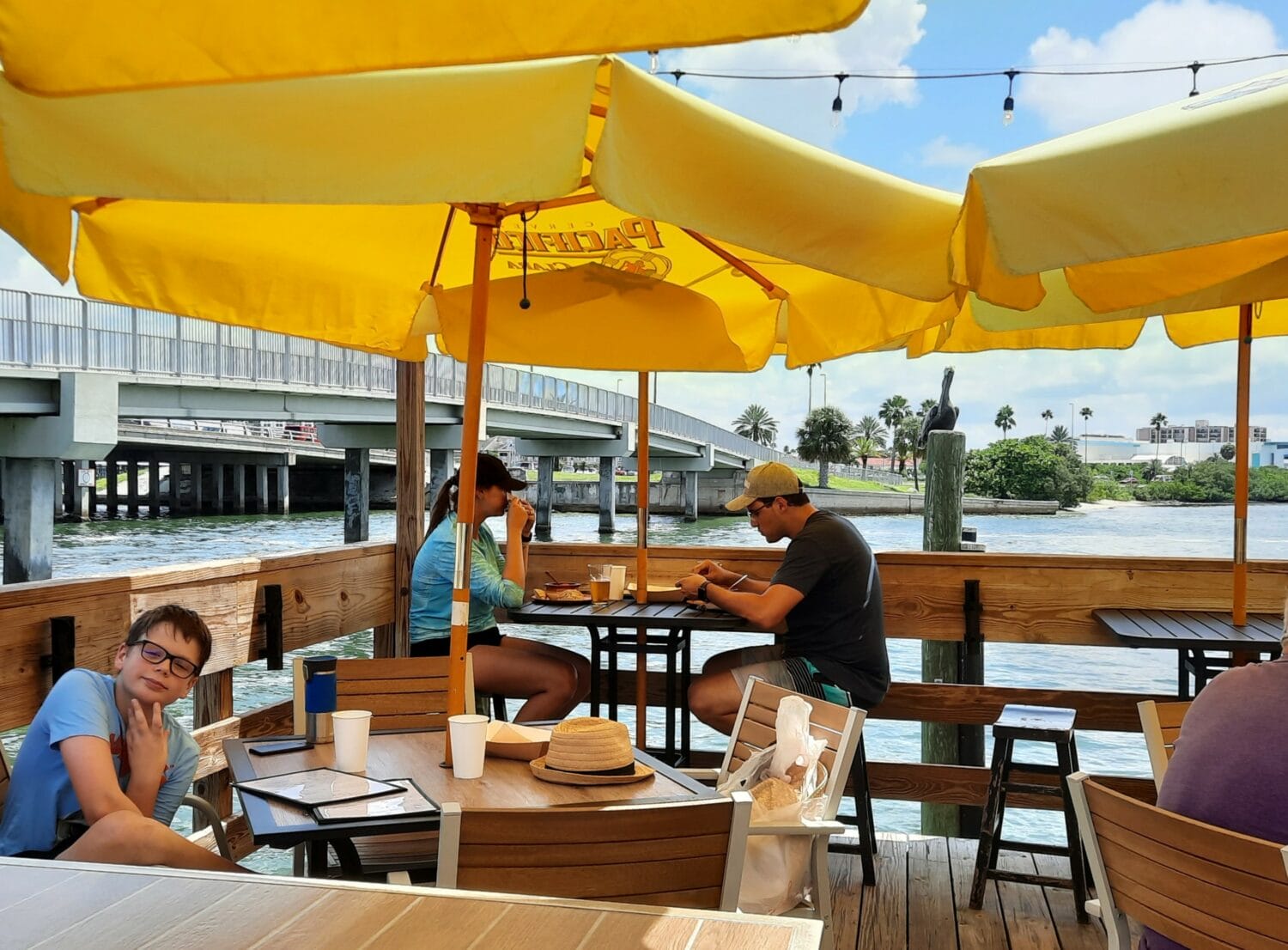 guests enjoying a meal under yellow umbrellas at a waterfront restaurant with a view of the bridge in the background