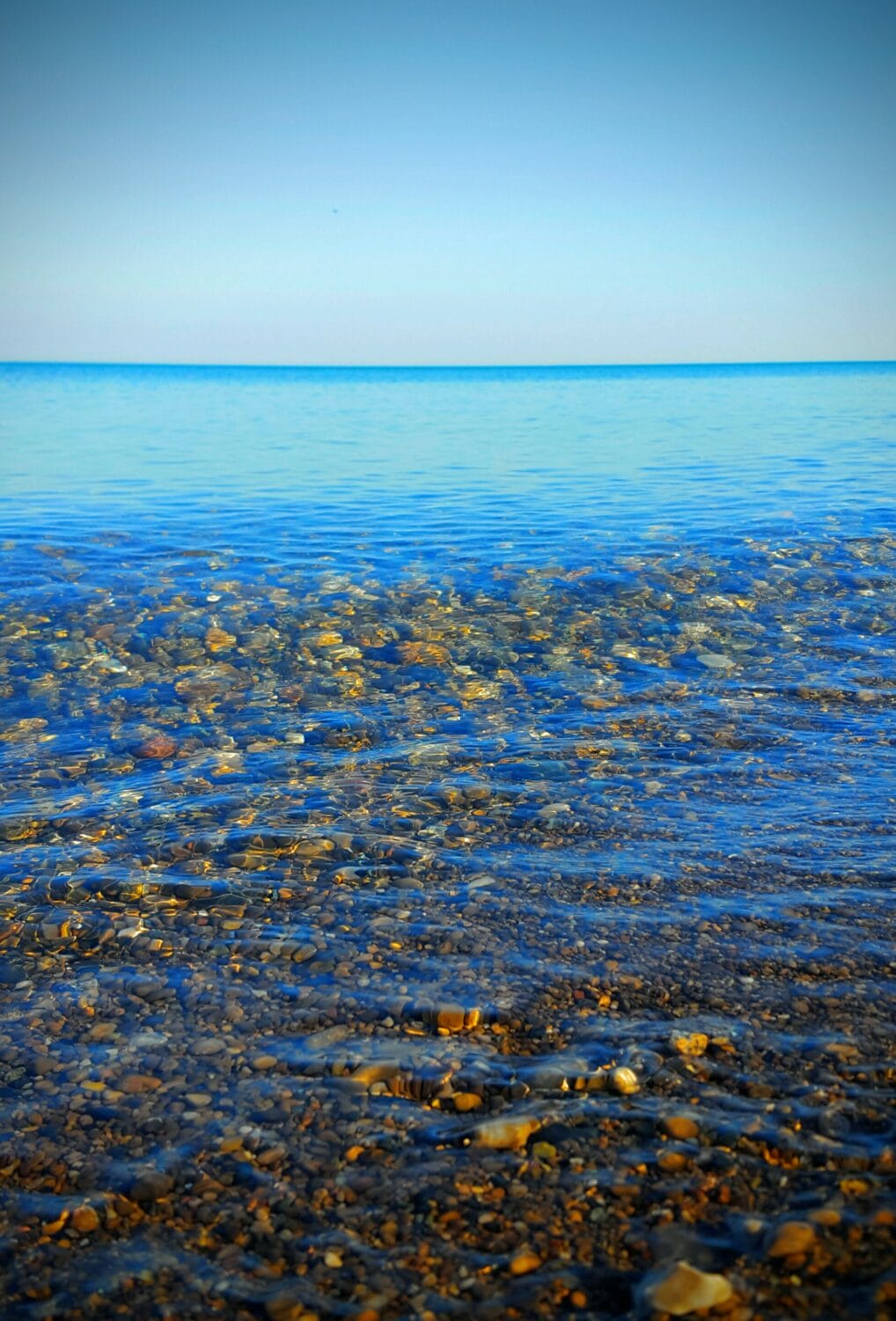 the clear waters of the beach gently lap over a pebble strewn shore under a clear blue sky