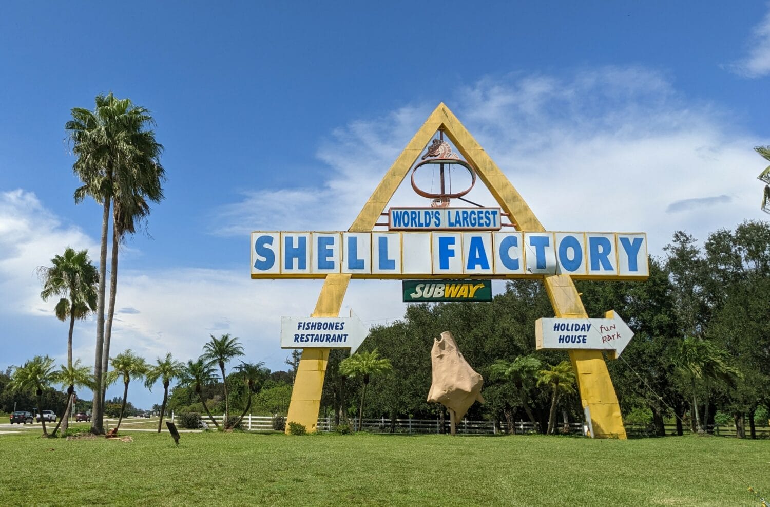 the entrance sign to the worlds largest shell factory featuring bold lettering and a tropical setting with palm trees