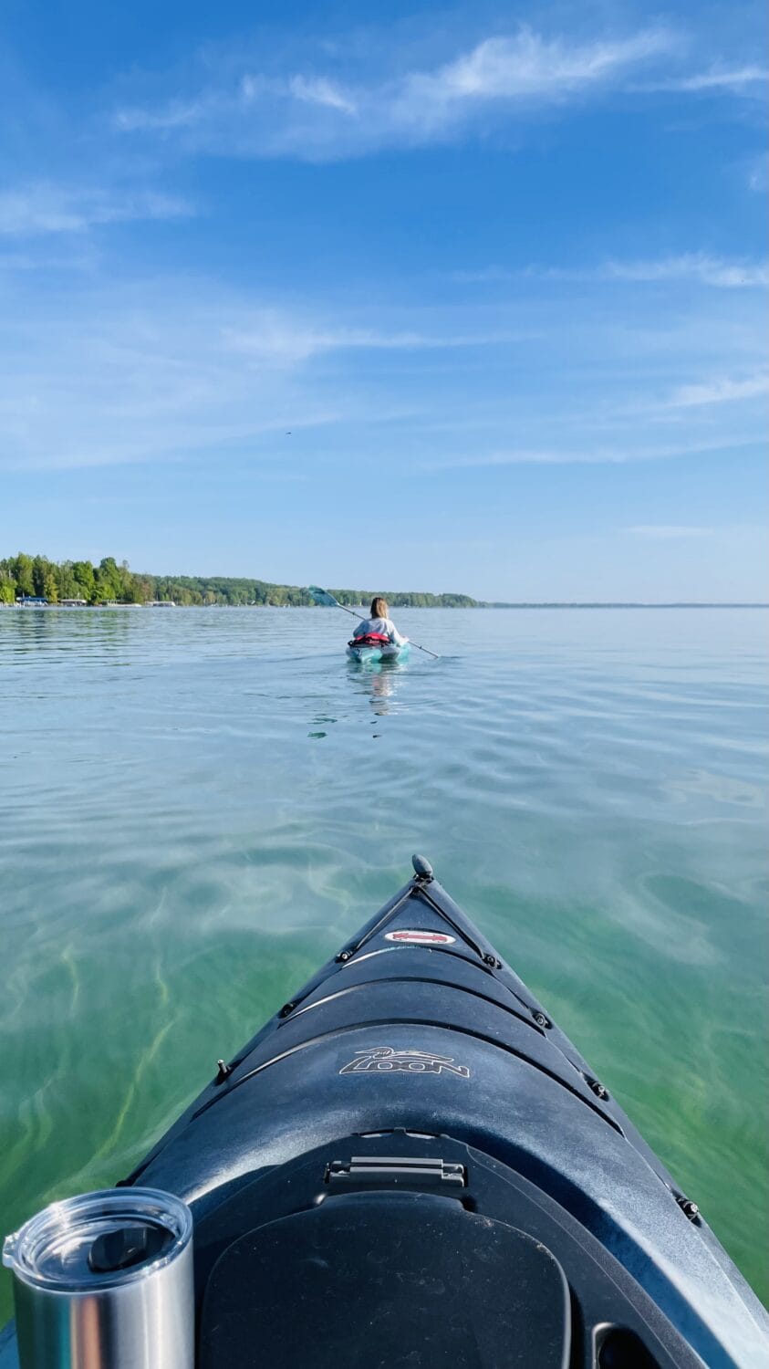 the view from a kayak on a clear lake with another kayaker in the distance against a backdrop of a blue sky