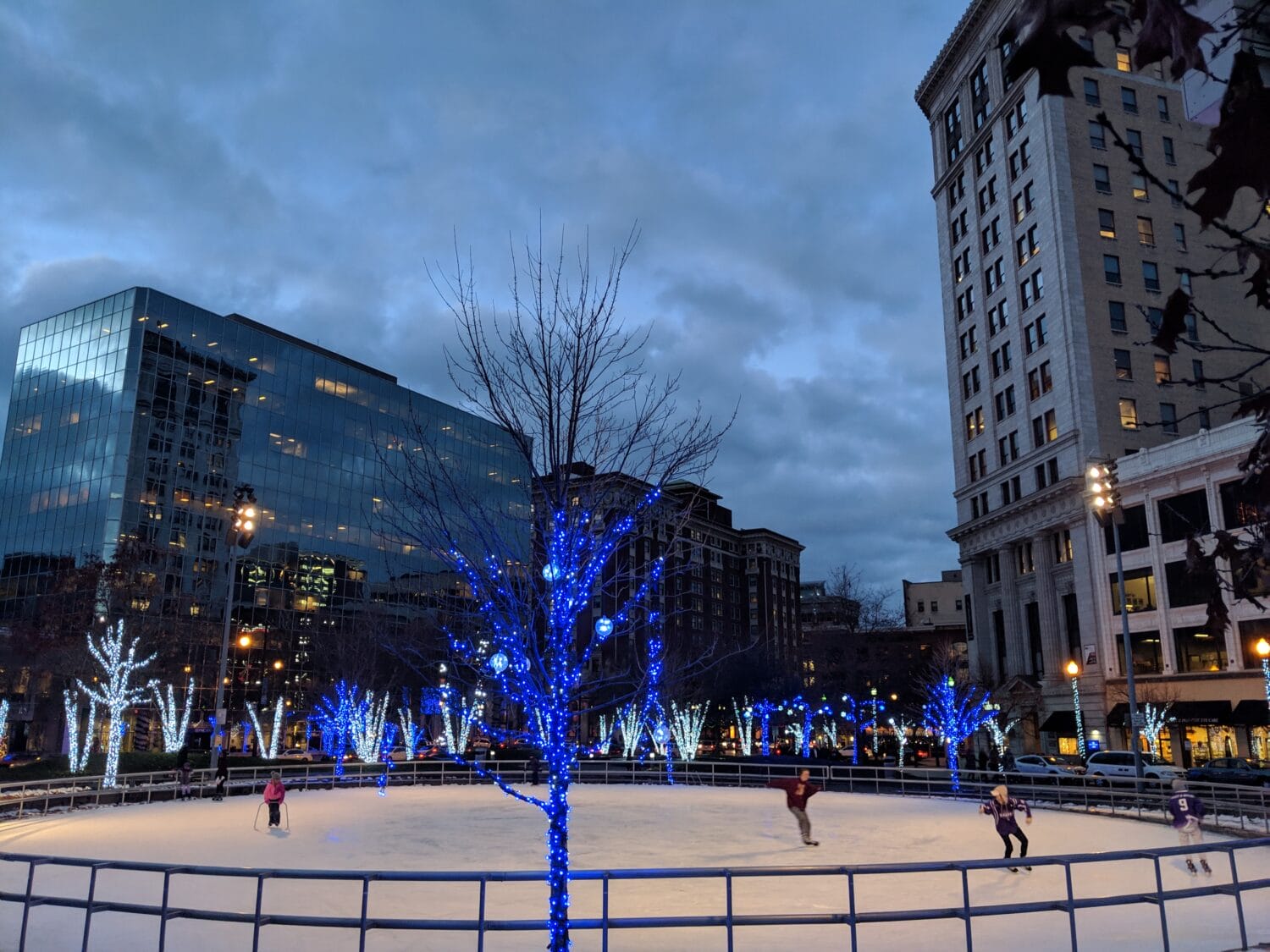 twilight descends on the lively outdoor ice skating rink framed by blue lit trees and modern city buildings