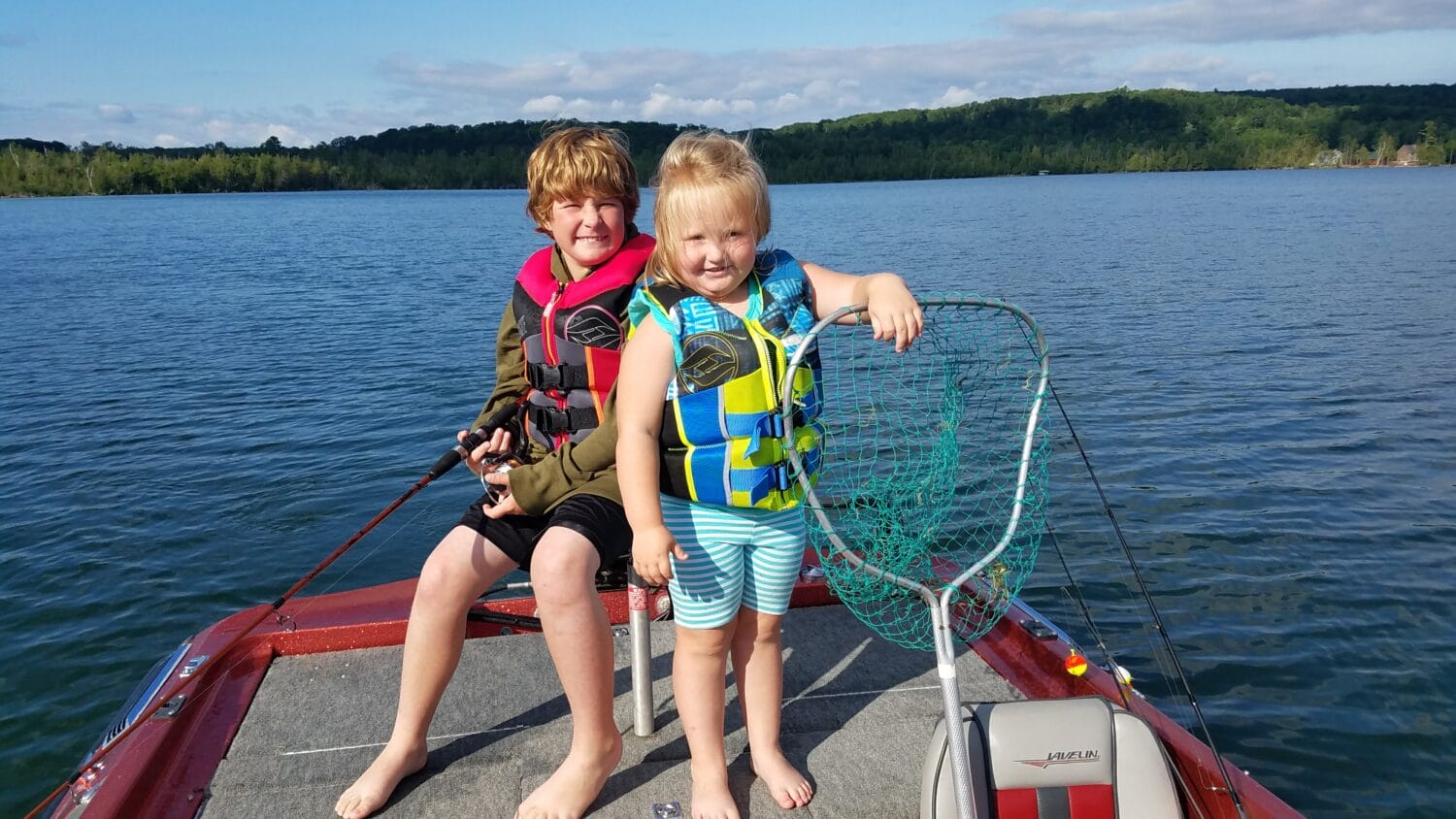 two children in life jackets standing on a boat ready for a day of fishing on a sunlit lake