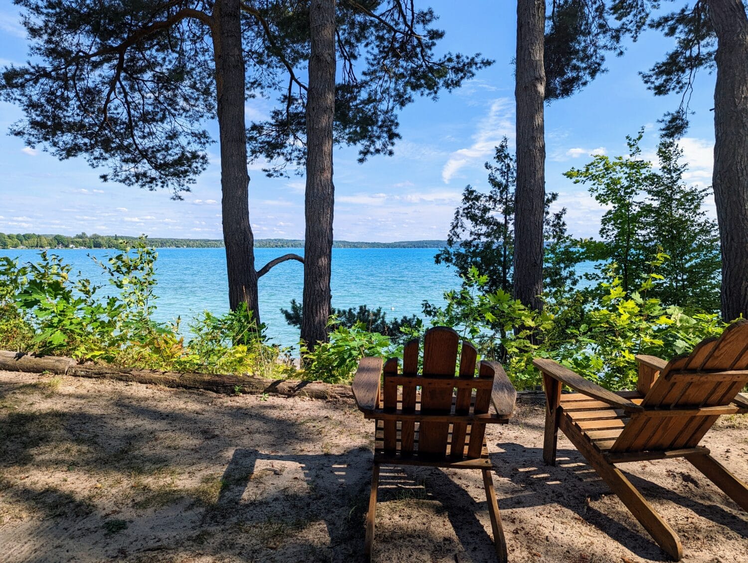 two wooden chairs facing a tranquil lake view from a shady sandy beach
