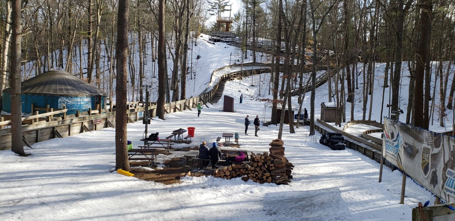 Visitors gather at the base of a snow-covered luge track, with a cozy seating area by a stack of wood and a view of the track winding through the trees.