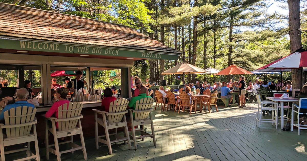 a bustling outdoor deck at Boone’s Long Lake Inn with guests enjoying meals and drinks under the shade of umbrellas, surrounded by tall pine trees.