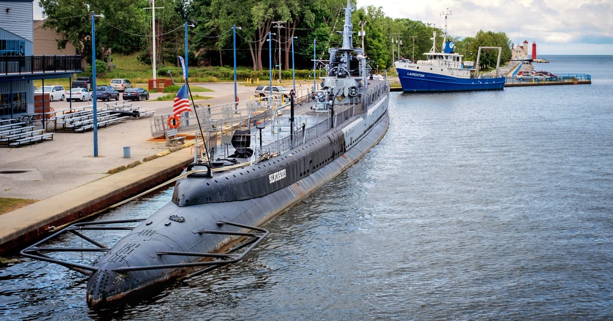 a historic submarine, the uss silversides submarine, moored in a calm marina, with another boat docked in the distance and a clear view of the shoreline and surrounding facilities.
