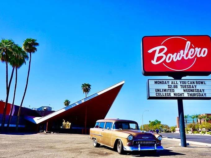 The iconic angular roofline and vintage car create a perfect time capsule of mid-century modern design, complete with swaying palm trees against the Arizona sky.