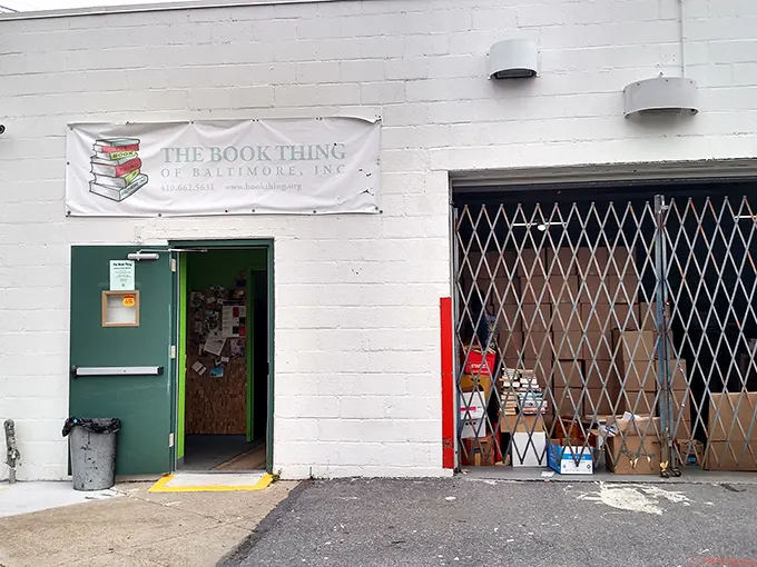 The unassuming exterior of literary heaven – a white cinder block building with a simple banner that promises bookish treasures beyond the green door.