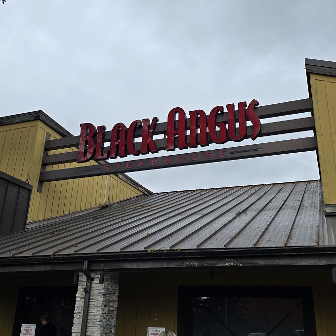 The glowing red signage of Black Angus beckons hungry travelers like a carnivorous lighthouse guiding ships to beefy salvation.