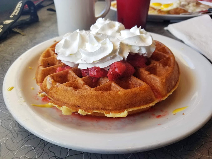 A waffle that understands the assignment – golden, crisp exterior giving way to fluffy interior, topped with strawberries and whipped cream. Breakfast or dessert? Yes.