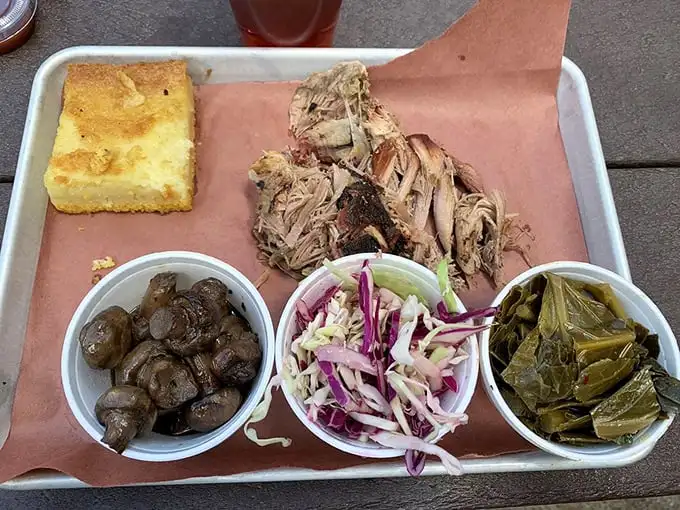 A metal tray of pulled pork perfection flanked by the holy trinity of barbecue sides: mushrooms, slaw, and collards. That cornbread isn't just a side—it's an essential tool.