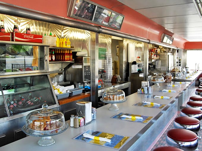 The counter where diner magic happens—chrome stools awaiting regulars, while pie domes protect treasures sweeter than Fort Knox. Decisions were never this delicious.