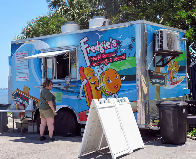 Paradise found: A cheerful blue food truck nestled among palm trees, where waterfront views come standard with every hot dog order.