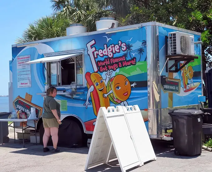 Paradise found: A cheerful blue food truck nestled among palm trees, where waterfront views come standard with every hot dog order.
