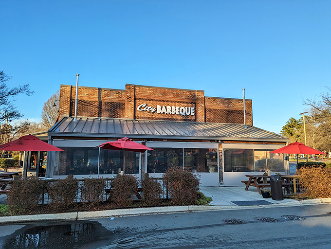 The brick fortress of flavor stands proud with its metal roof and welcoming red umbrellas—a barbecue beacon calling hungry travelers home. 