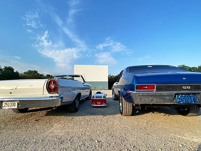 Classic American cars flank a tiny pedal car, three generations of automotive history facing the silver screen—nostalgia in its purest form.