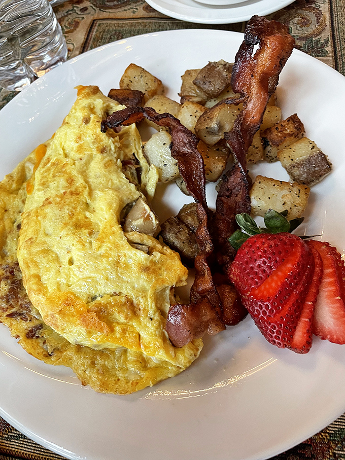Morning masterpiece: A perfectly crafted omelet alongside crispy bacon, seasoned potatoes, and fresh strawberries – breakfast that makes you forget to check your phone.