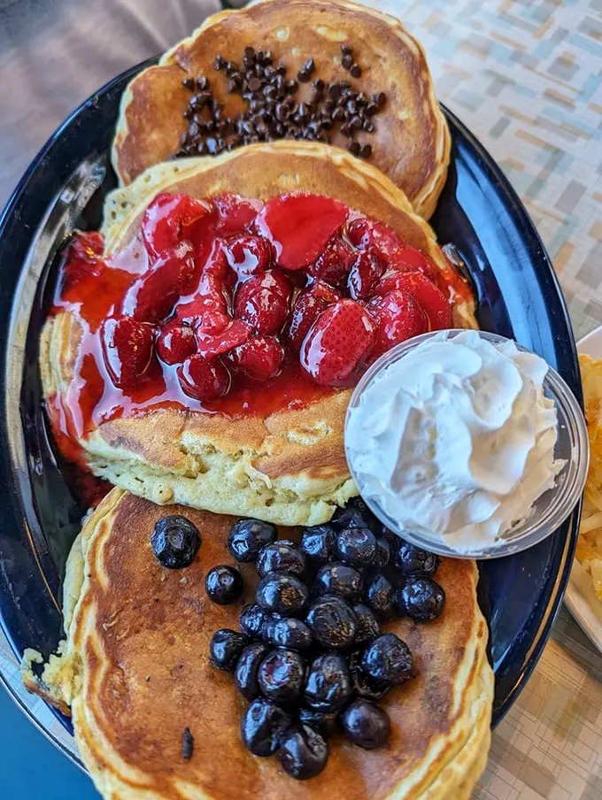 Pancake perfection in triplicate: chocolate chip, strawberry, and blueberry create the holy trinity of breakfast indulgence. That whipped cream isn't just a garnish—it's the exclamation point!