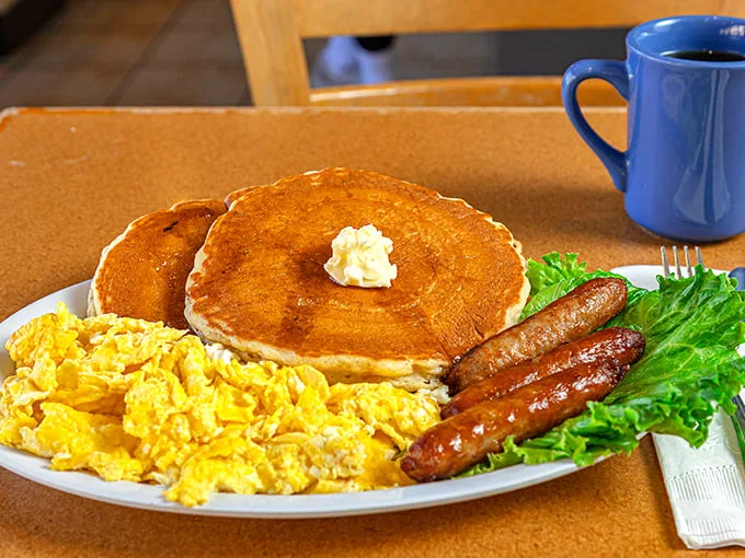 This hearty breakfast platter features fluffy scrambled eggs, golden pancakes topped with butter, savory sausage links, and a fresh garnish, served with a hot cup of coffee.