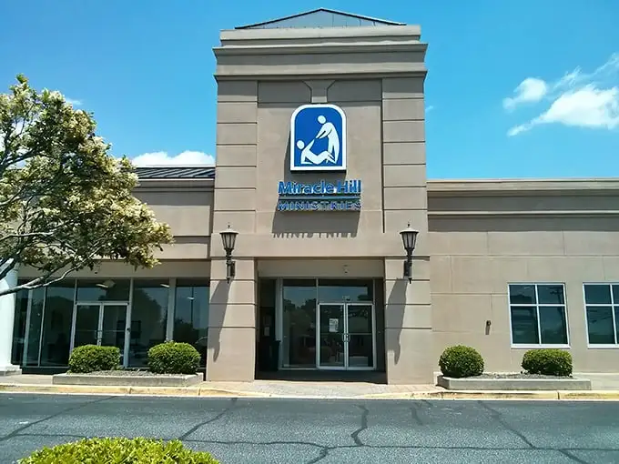 The iconic blue signage of Miracle Hill Thrift Store stands out against the beige building like a beacon for bargain hunters across Greenville.