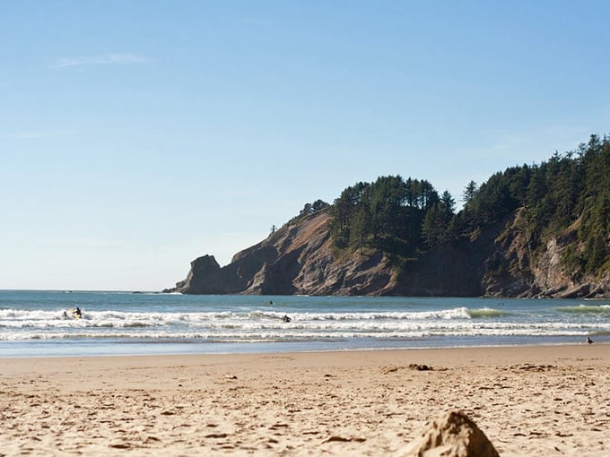 Nature's perfect postcard – dramatic headlands embrace golden sands while surfers dance on gentle waves beneath an impossibly blue Oregon sky.