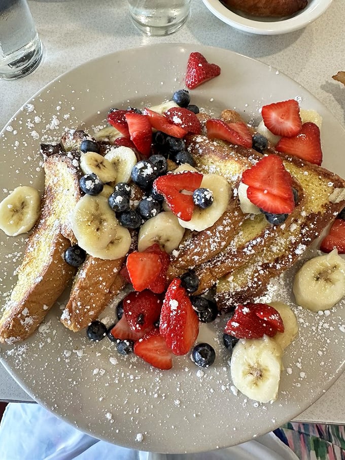French toast that's having an identity crisis in the best possible way—crowned with a berry and banana carnival and enough powdered sugar for snow angels.
